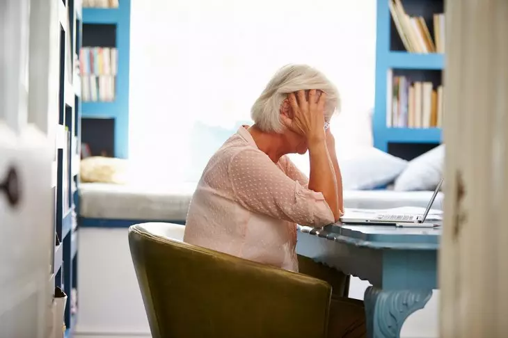 Een gestresste oudere vrouw leunt over haar laptop en bureau in een thuiskantoor